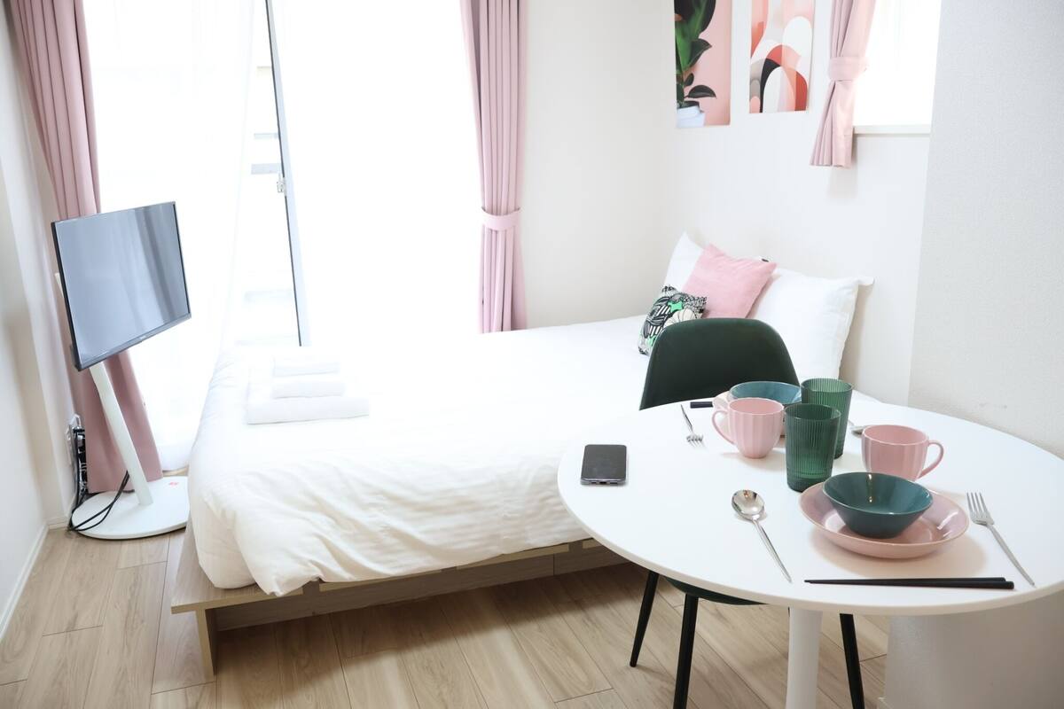Quarto de hotel bem arrumado com uma cama, uma TV, uma mesa de jantar com chávenas e uma tigela, e uma janela com cortinas.