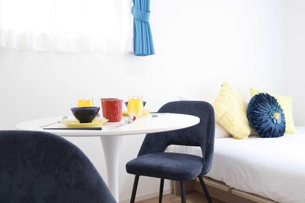 Quarto moderno com uma cama branca, uma mesa de jantar redonda com cadeiras e uma poltrona azul.