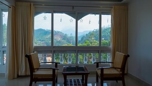 Quarto com duas cadeiras e uma mesa, janelas amplas com cortinas, e vista para uma paisagem montanhosa.