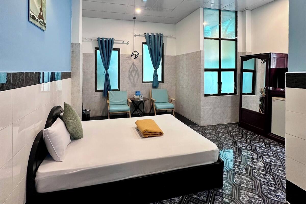 Quarto de hotel com cama, duas cadeiras, uma mesinha e janela com cortinas.