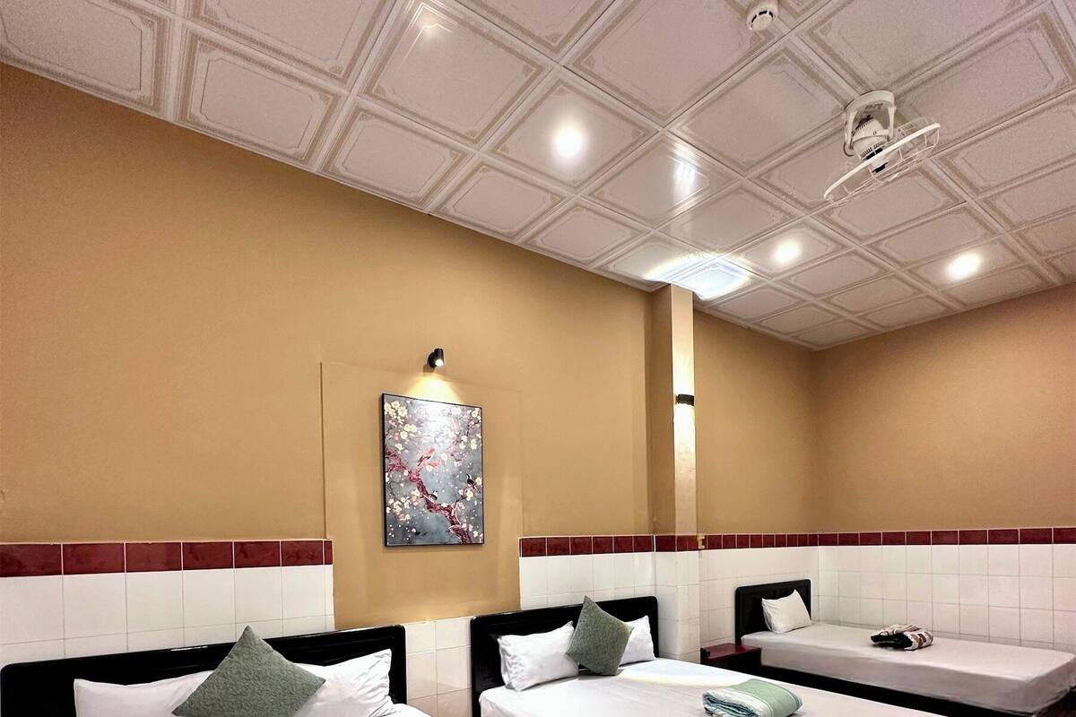 Quarto de hotel com três camas individuais, cada uma com uma almofada e uma toalha dobrada. O quarto possui piso de azulejo e teto branco com iluminação embutida.
