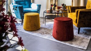 Uma sala de estar moderna com uma poltrona azul, um banquinho amarelo e um banquinho vermelho. Há um espelho grande, um abajur e uma janela com cortinas.