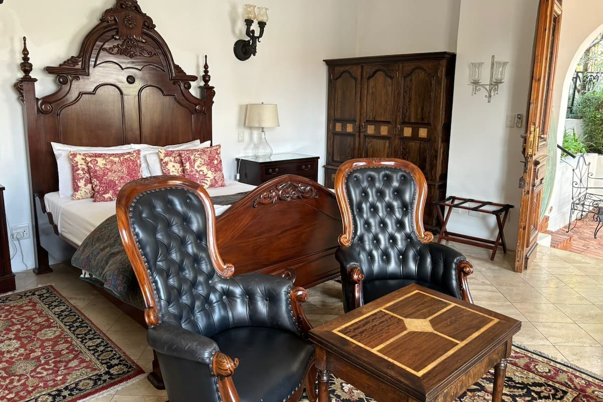 Um quarto com uma cama de madeira grande, duas poltronas estofadas, uma mesa de madeira pequena e uma mesa de cabeceira com abajur.