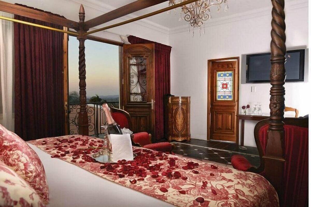 Uma cama com dossel adornada com pétalas de rosa, uma escrivaninha de madeira, uma janela grande e um candelabro.
