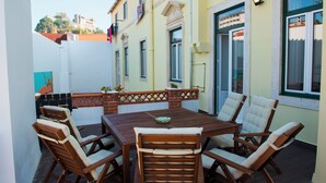 Um pátio ao ar livre com mesa de madeira e seis cadeiras, um pequeno jardim e vista para um edifício histórico ao fundo.