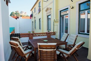 Um pátio ao ar livre com mesa de madeira e seis cadeiras, um pequeno jardim e vista para um edifício histórico ao fundo.