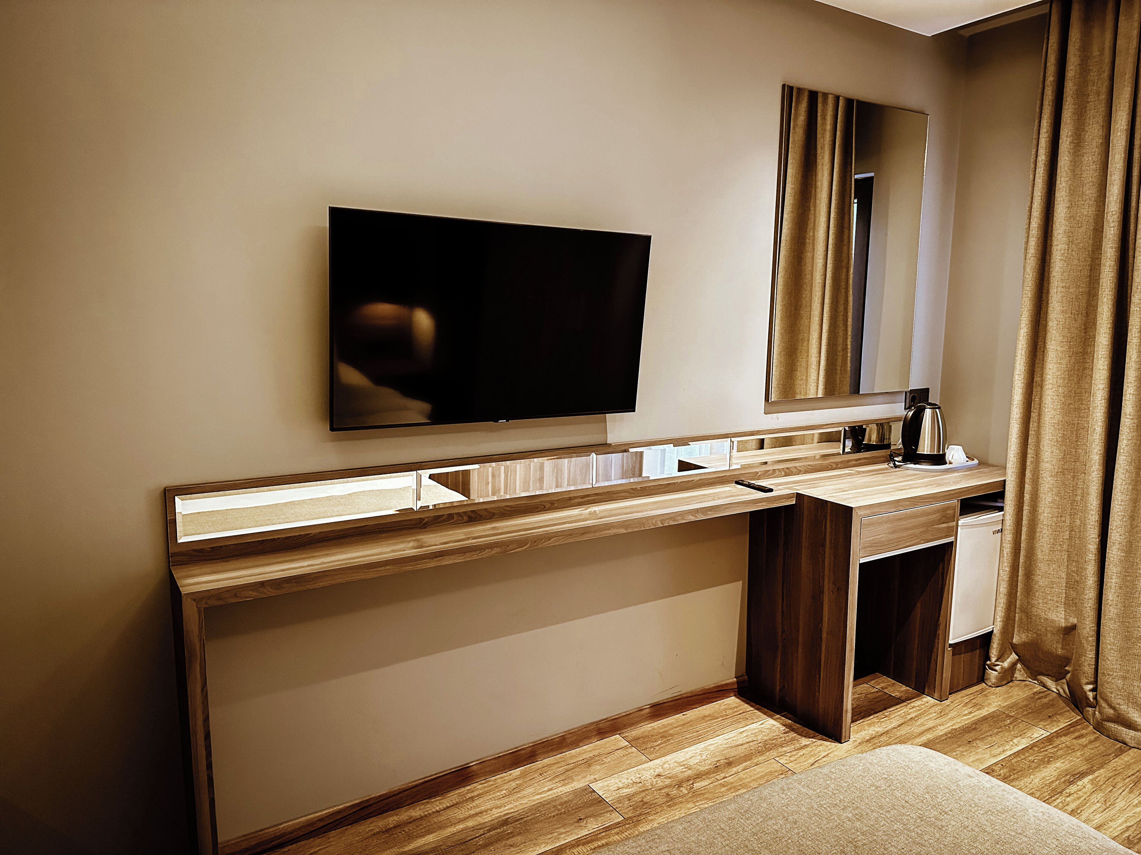 Quarto de hotel moderno com televisão de tela plana montada acima de uma toucador de madeira, um espelho e uma cafeteira pequena.