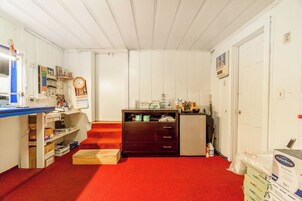 Quarto com carpete vermelho, paredes brancas, escrivaninha de madeira, pequeno frigorífico e cómoda de cor escura.