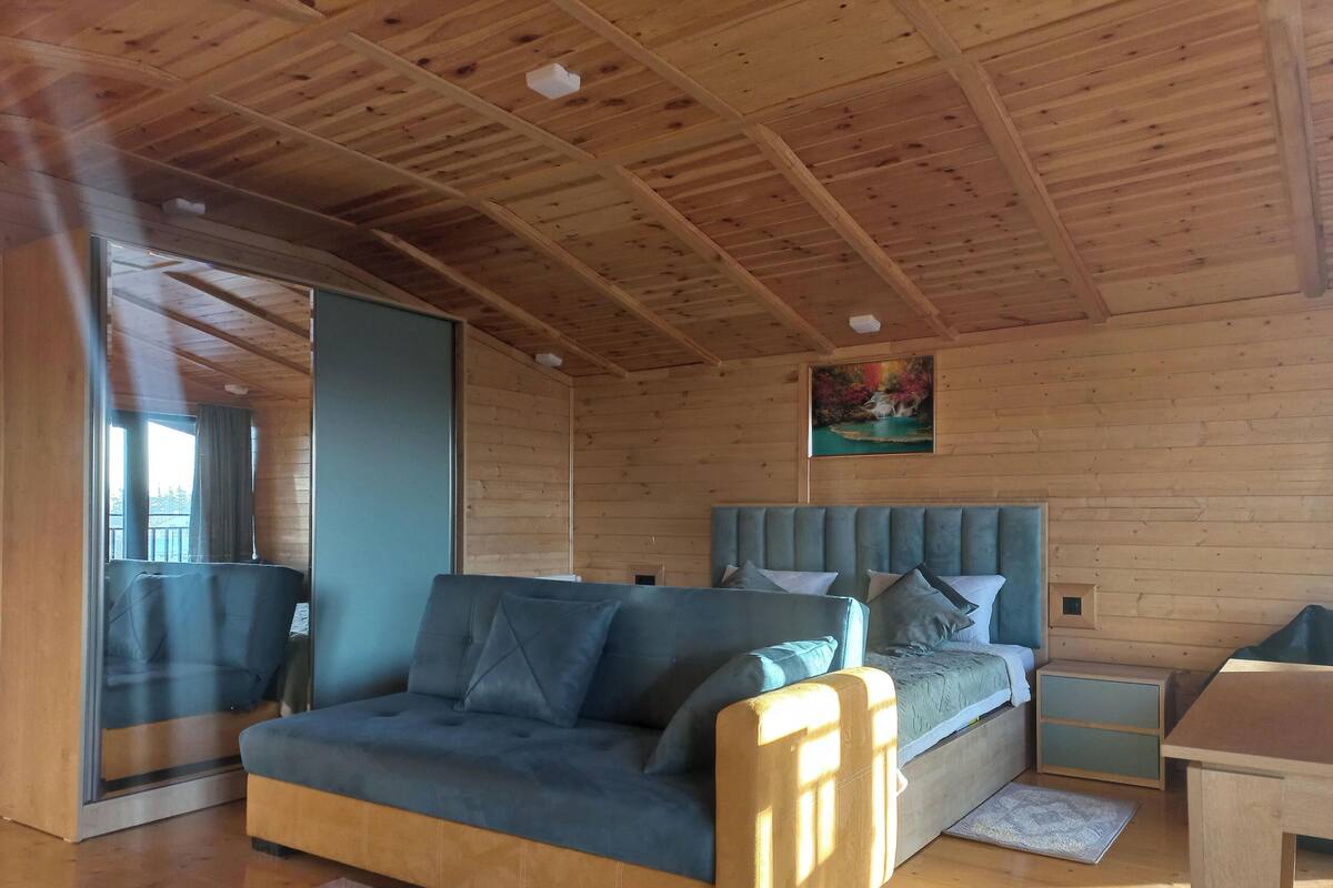 Interior de uma cabana de madeira com sofá, cama com encosto, uma mesinha e um quadro na parede.