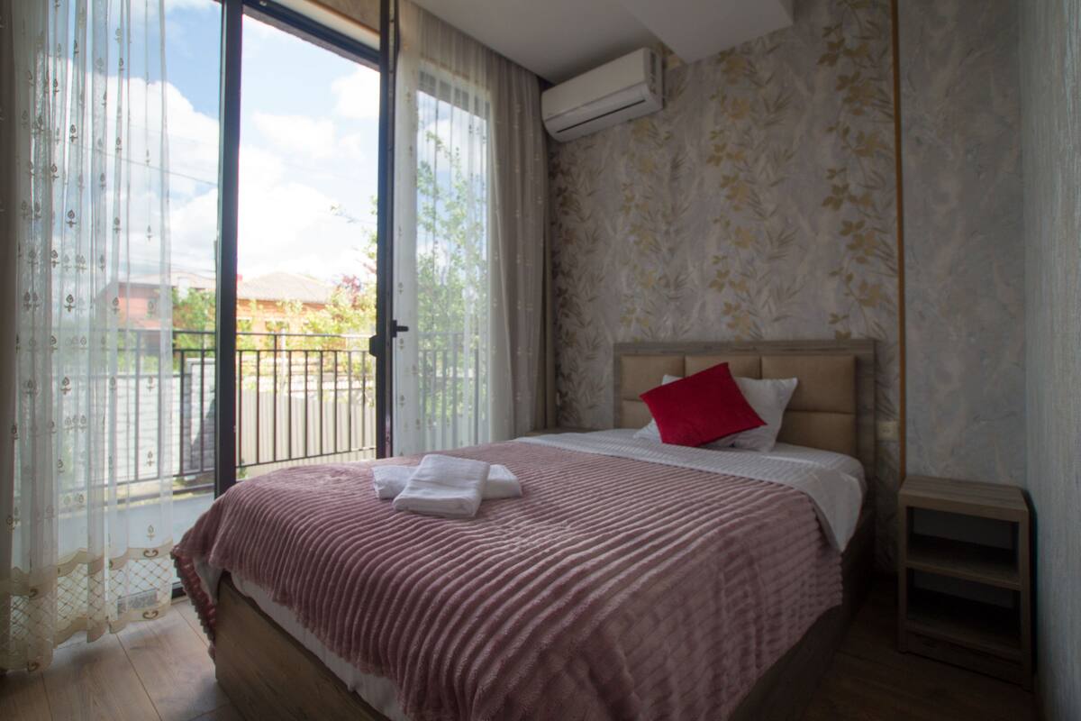 Quarto com cama, almofada vermelha e janela com vista para o exterior.