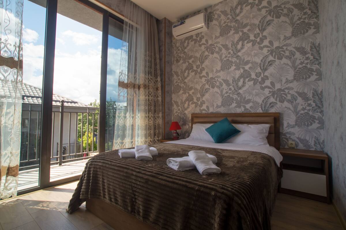 Quarto de hotel com uma cama grande, uma mesa de cabeceira, um abajur vermelho e vista para o exterior.