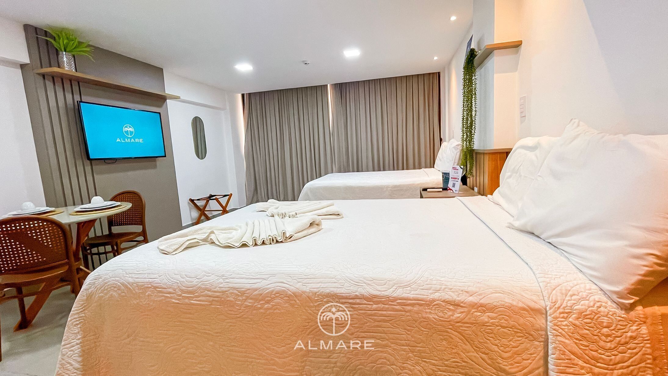 Quarto de hotel com cama, escrivaninha, cadeira, televisão e planta.
