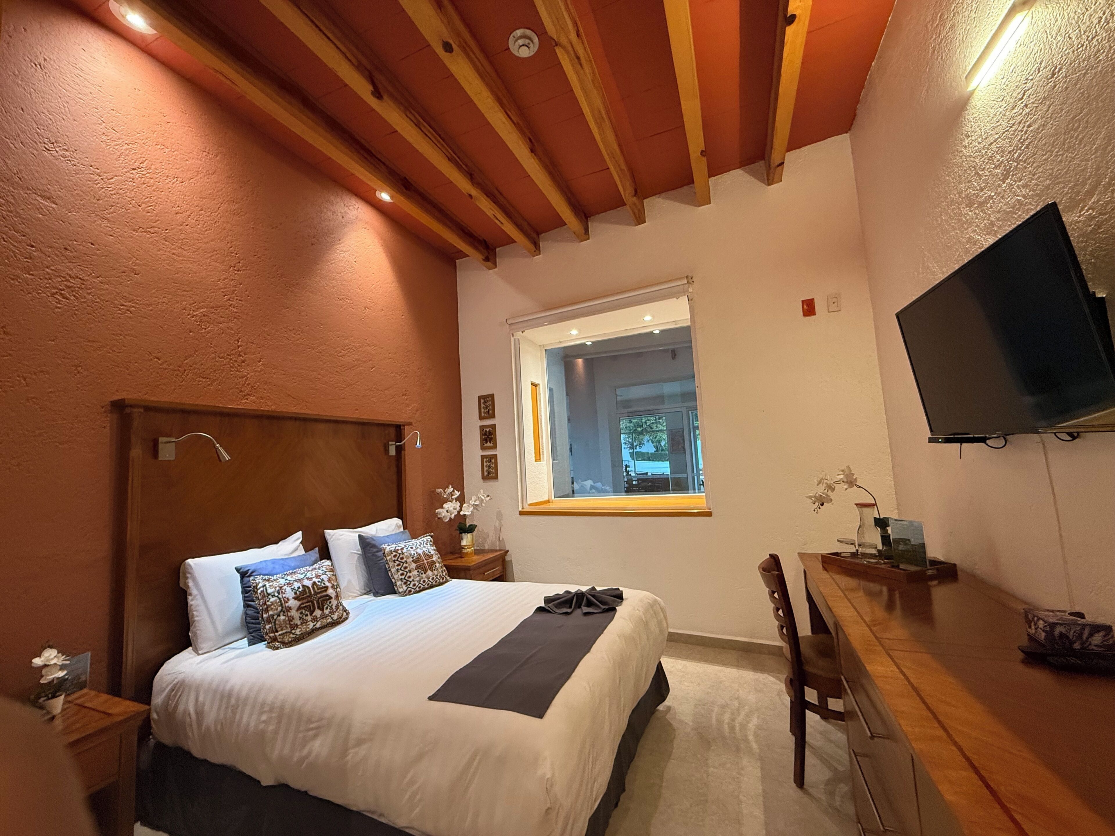 Quarto com teto de madeira, cama com roupa de cama branca e almofadas estampadas, mesa de cabeceira com um vaso de flores, janela com vista para o exterior e televisão de ecrã plano na parede.