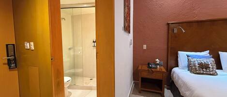 Quarto de hotel com porta de madeira, box de vidro no chuveiro, mesa de cabeceira com telefone e cama com almofadas.