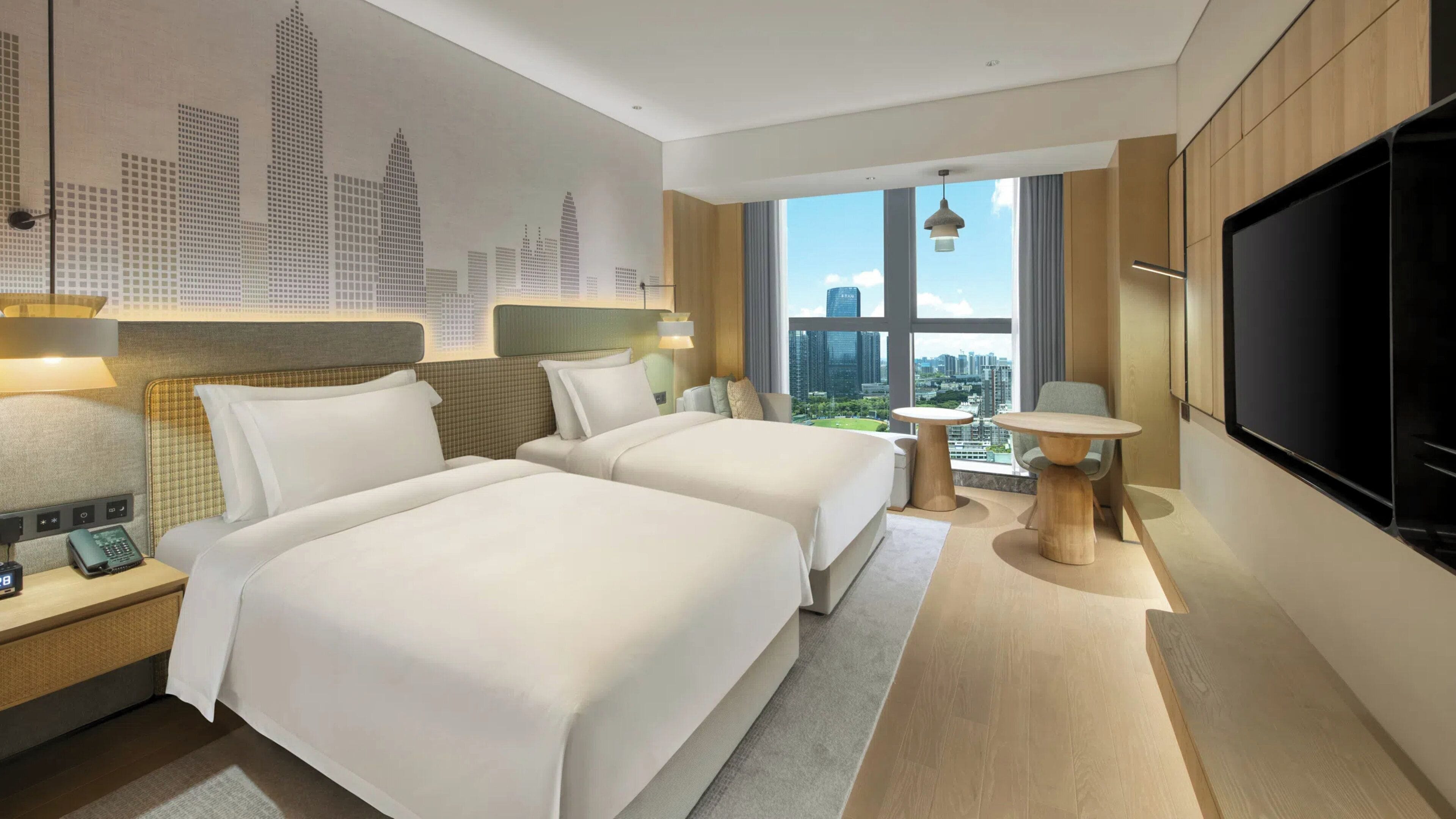 Quarto de hotel moderno com duas camas, uma televisão grande e vista para a cidade.
