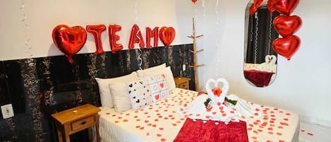 Quarto com cama, almofadas, mesa de cabeceira, espelho e balões em formato de coração.