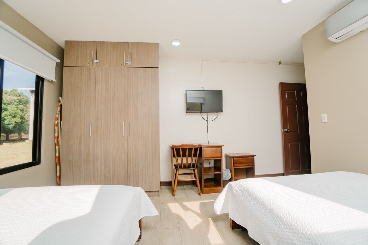 Quarto de hotel com duas camas, um guarda-roupa de madeira, uma mesa com cadeira, televisão e uma porta.