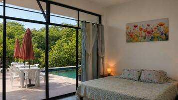 Quarto com cama, um quadro e vista para uma piscina e vegetação lá fora.