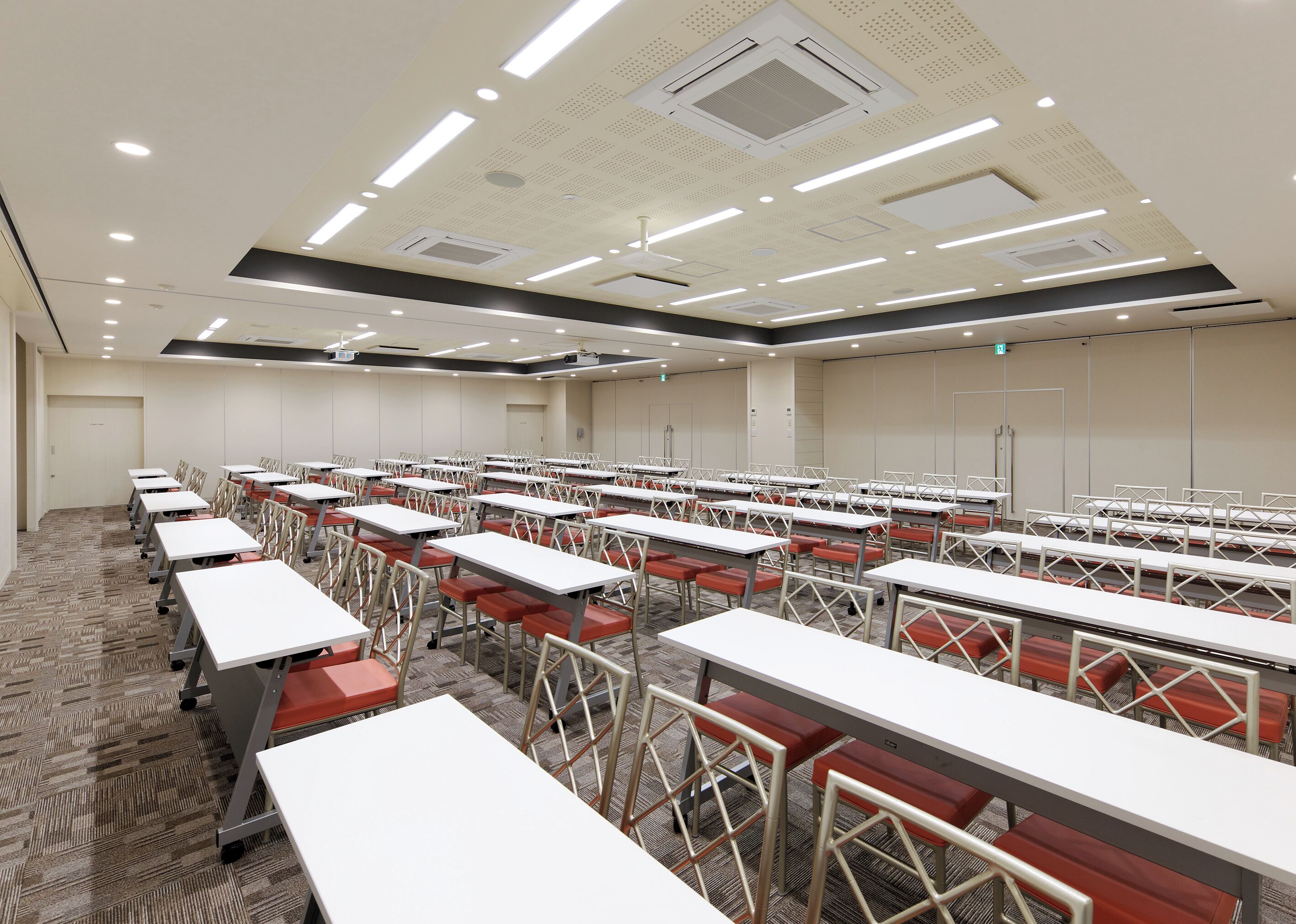 Sala de conferências com fileiras de mesas e cadeiras, equipada com iluminação no teto e ventilação.