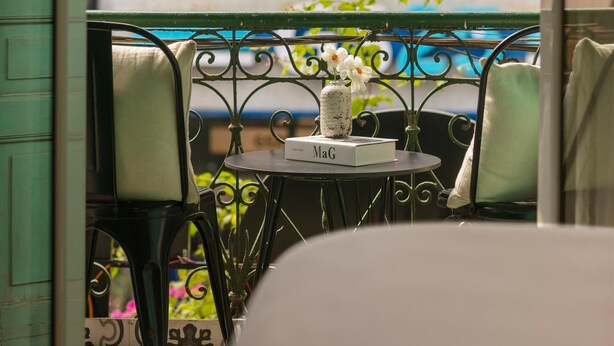 Um café com mesas ao ar livre, uma mesa com um vaso e uma caixa com a etiqueta 'MaG', e uma fachada com o letreiro 'CAFE25'.