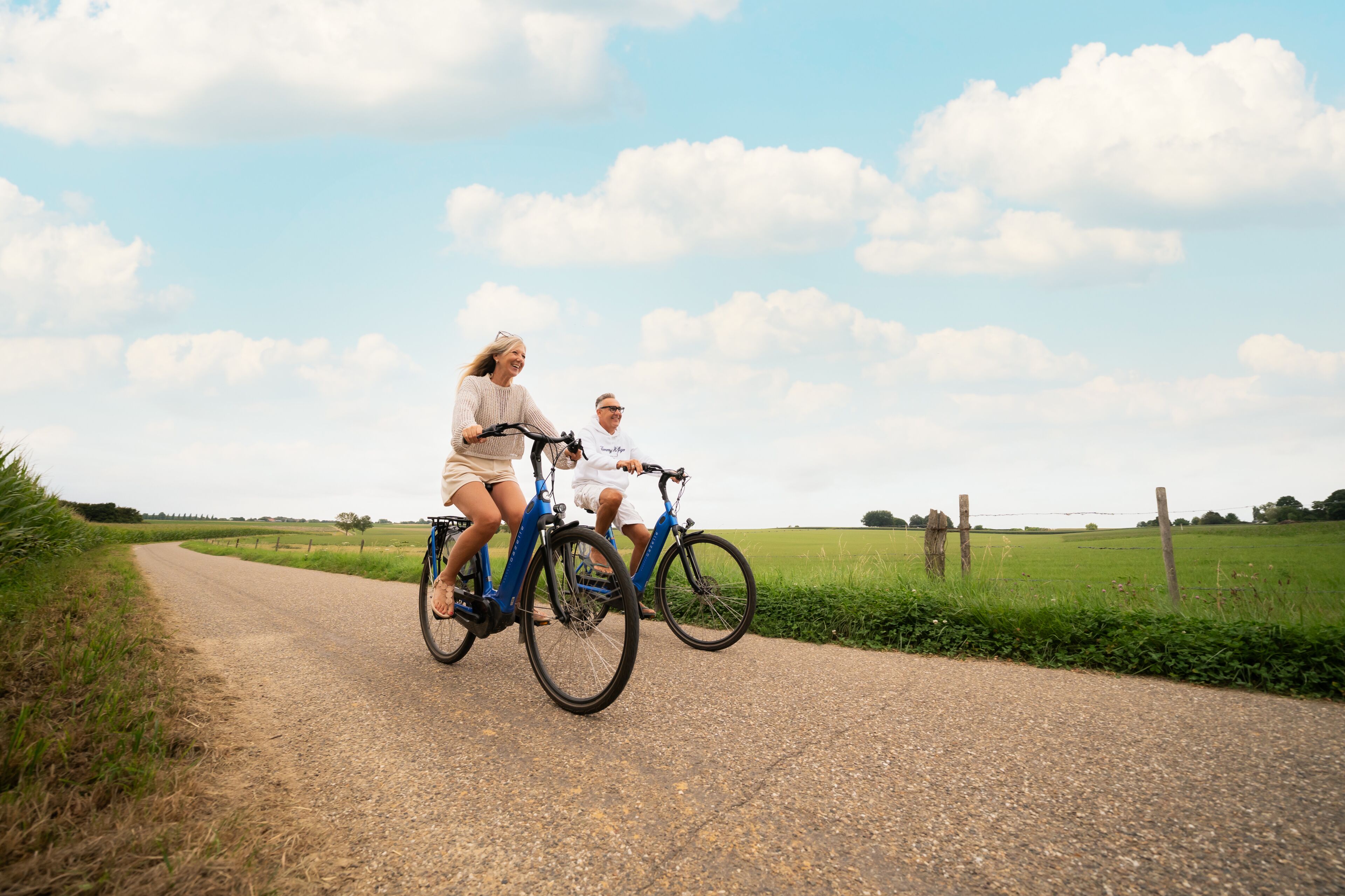 Duas pessoas em bicicletas numa estrada rural, com campos abertos e céu limpo.