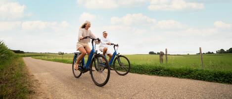 Duas pessoas em bicicletas numa estrada rural, com campos abertos e céu limpo.
