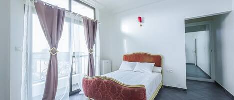 Quarto com cama grande, janela com cortinas, tapete pequeno e luminária vermelha fixada na parede.