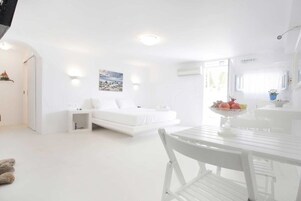 Um quarto claro com uma cama, uma mesa pequena com frutas e uma cadeira branca.