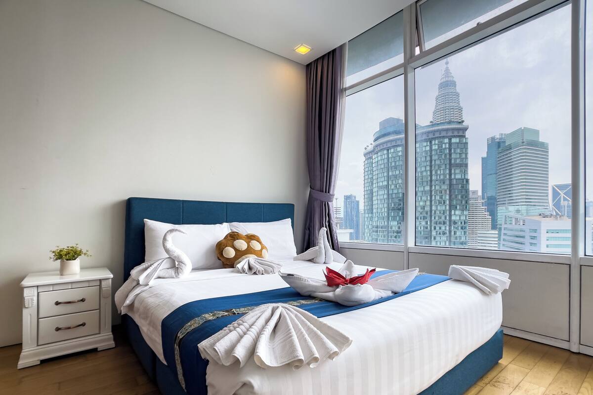 Quarto de hotel com uma cama grande, mesa de cabeceira, planta em vaso e vista para edifícios da cidade.