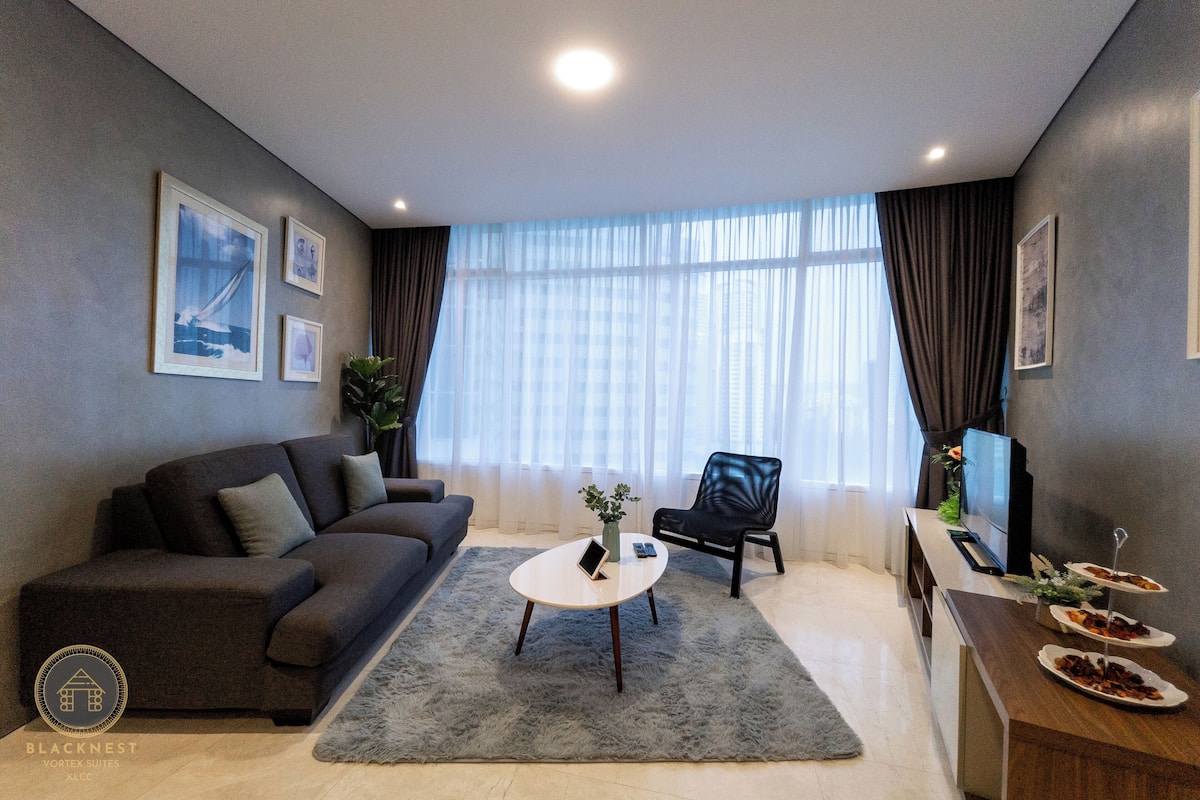 Uma sala de estar moderna com um sofá cinza, uma mesa de centro redonda, uma poltrona de couro preto e uma TV sobre um suporte de madeira.