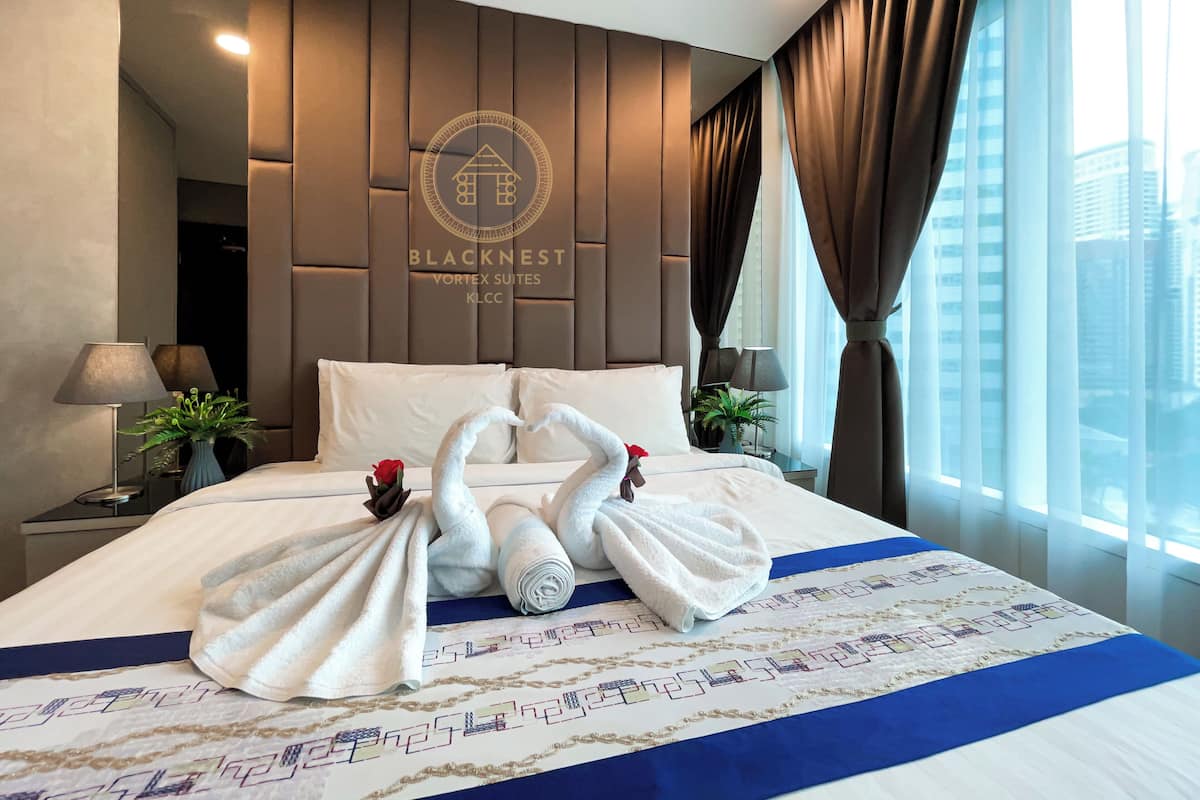 Quarto de hotel com uma cama grande, cabeceira com logotipo, janela com cortinas e mesa de cabeceira com abajur e planta.