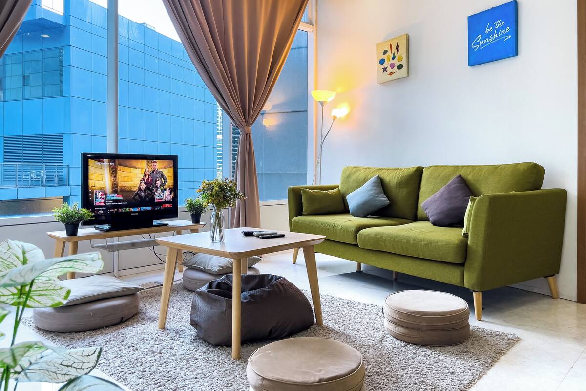Uma sala de estar moderna com um sofá verde, uma TV de tela plana, uma mesa de centro e uma janela grande com cortinas.