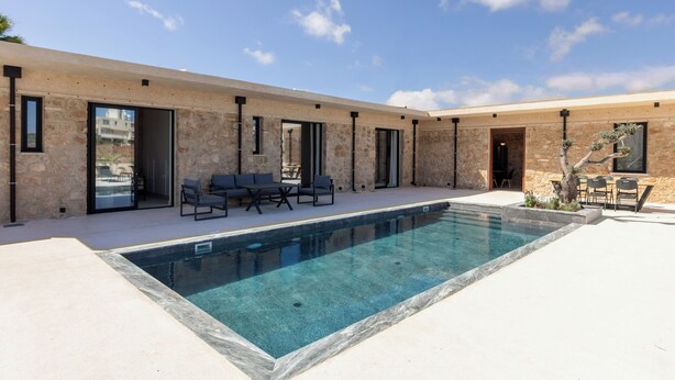 Uma área de piscina moderna ao ar livre com fachada de pedra, assentos externos e céu azul límpido.