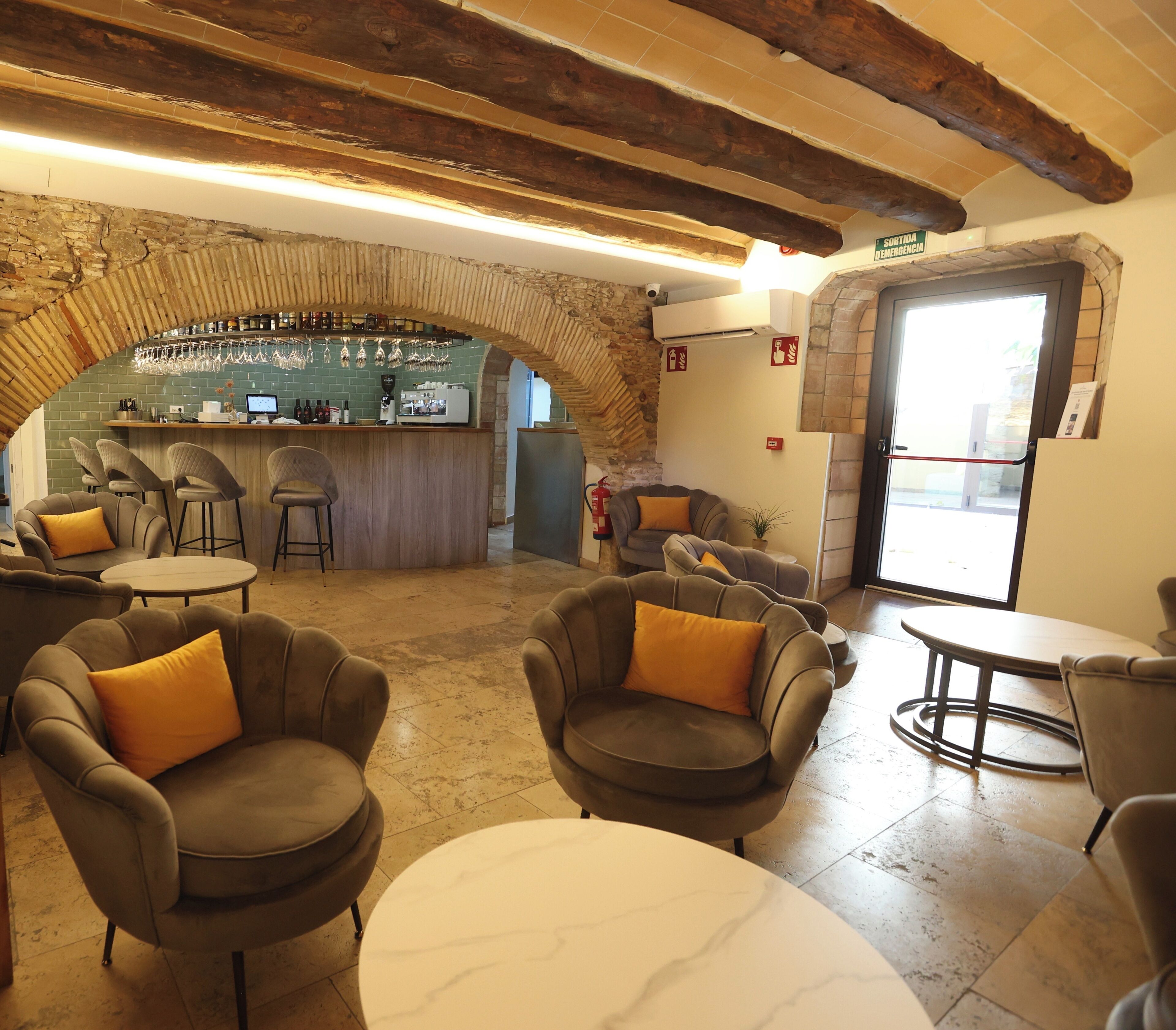 Uma área de bar com banquetas, um balcão e um extintor de incêndio. Cadeiras estofadas e uma mesa estão em primeiro plano.