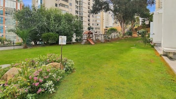 Uma área ajardinada bem cuidada com um parquinho, cercada por edifícios residenciais e árvores.