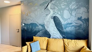 Um quarto com um grande mural na parede retratando um pássaro e árvores, um sofá com almofadas, e uma porta.