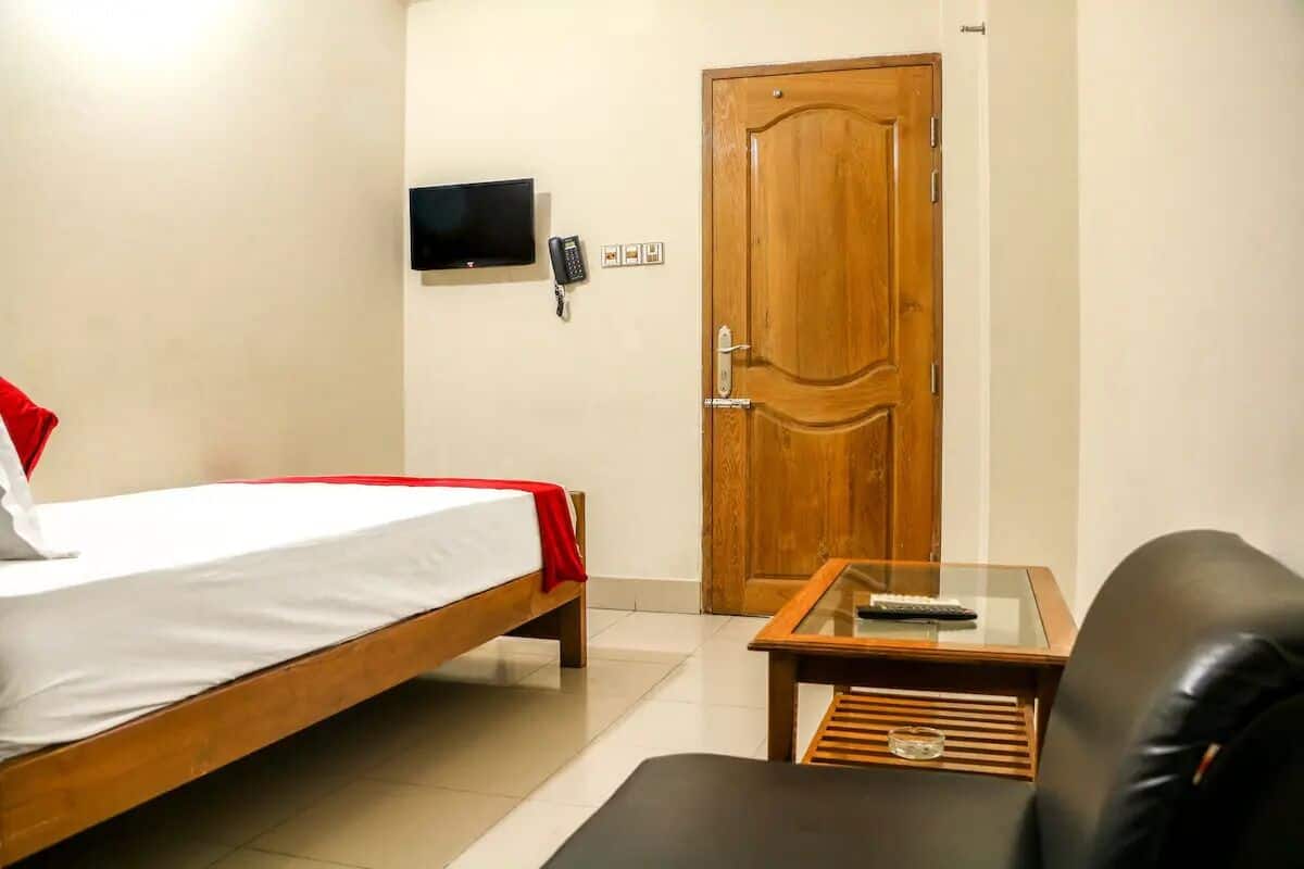 Quarto de hotel com cama, televisão fixada na parede, porta de madeira e uma pequena mesa com um controle remoto.