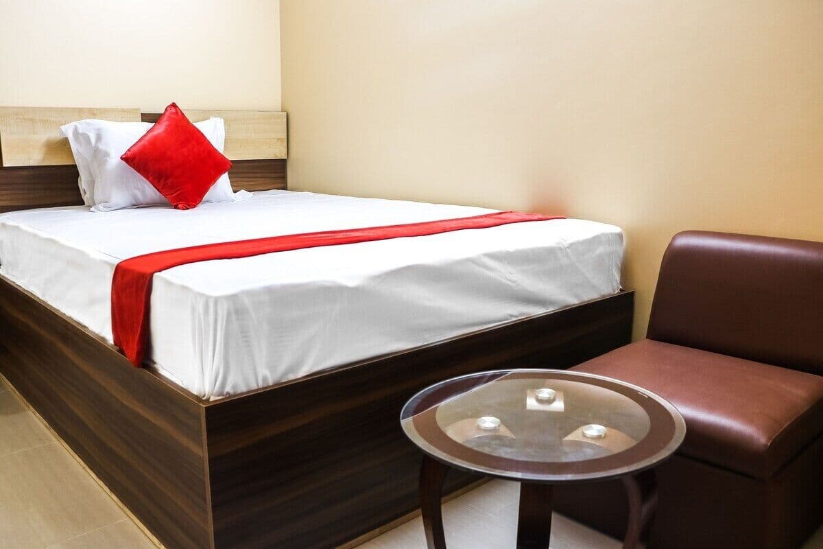 Quarto de hotel com uma cama, um cobertor vermelho, uma poltrona de couro marrom e uma mesa de vidro.