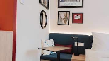 Quarto de hotel com uma cama, uma escrivaninha, uma cadeira e uma mesa pequena. Há quadros nas paredes e o chão é coberto por carpete.