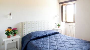 Um quarto com teto de madeira, uma cama com colcha azul, uma mesa de cabeceira com duas plantas em vasos e uma janela com vista.