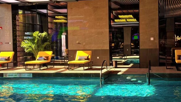 Uma área à beira da piscina com cadeiras de descanso amarelas, uma piscina cristalina e um letreiro neon com a inscrição 'THOUGHTS BECOME THINGS'.