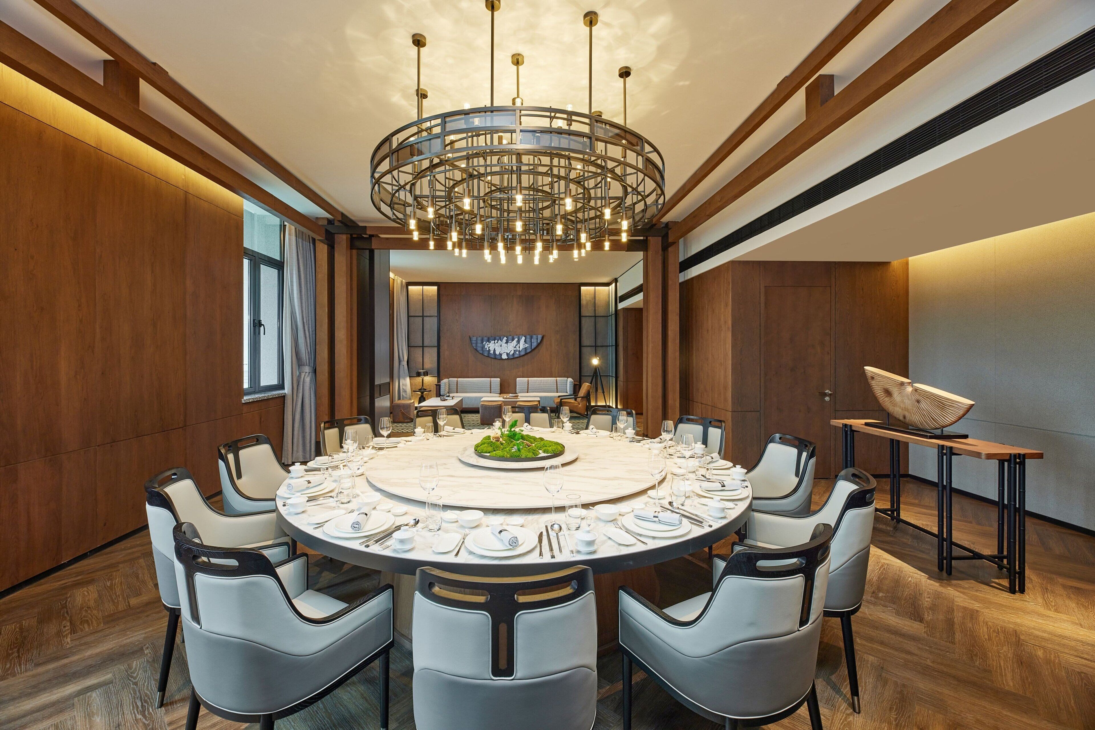 Uma sala de jantar moderna com uma mesa redonda posta para refeição, rodeada por cadeiras, e um lustre central.