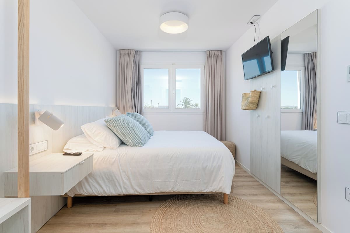 Quarto moderno com cama, mesa de cabeceira, abajur, televisão e um espelho grande.