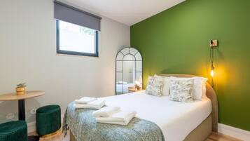 Quarto com cama, uma mesinha redonda com uma planta, um banquinho verde, janela com cortina e luminária fixada na parede.