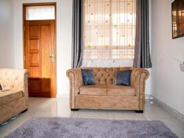 Uma sala de estar com um sofá bege, uma poltrona menor, um tapete macio, uma porta de madeira e uma janela com cortinas.