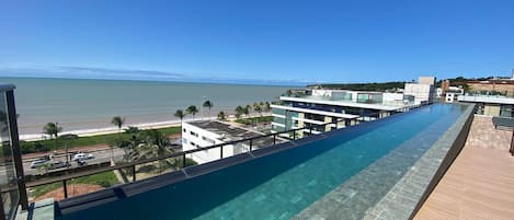 Uma piscina comprida com água cristalina, adjacente a um edifício moderno com vista para a praia e o oceano.