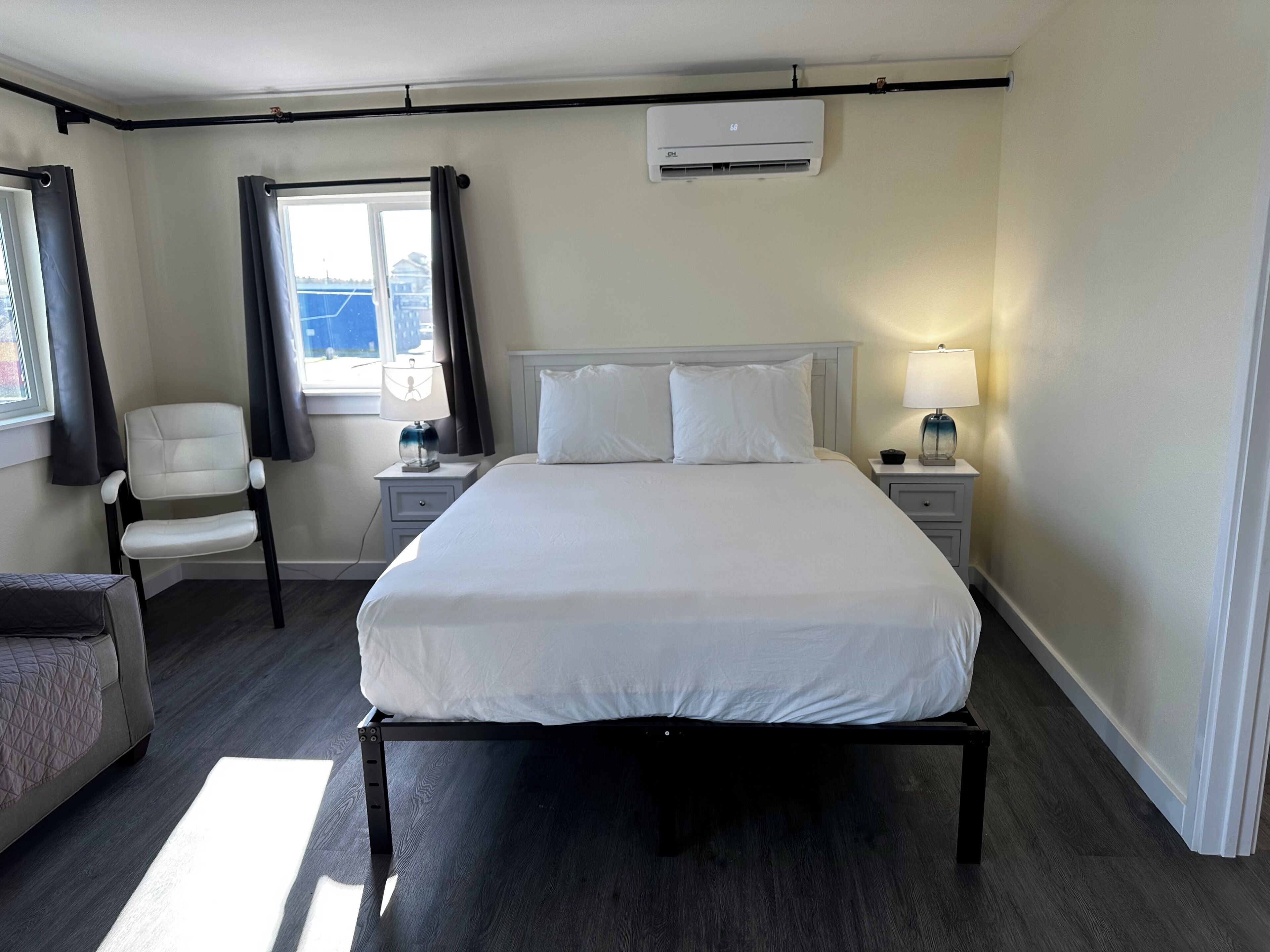 Quarto de hotel com uma cama grande, duas mesas de cabeceira com luminárias, uma cadeira, uma janela com cortinas e um ar condicionado.