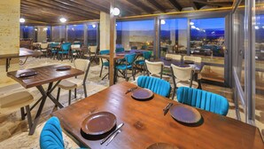 Um restaurante com mesas e cadeiras de madeira, janelas amplas e vista para a cidade à noite.