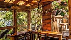 Uma zona de refeições rústica de madeira com um banco e cadeiras, rodeada por vegetação natural e uma estrutura de madeira com telhado inclinado.