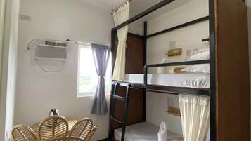 Quarto de dormitório com beliches, uma mesinha, uma cadeira e uma janela com cortinas.